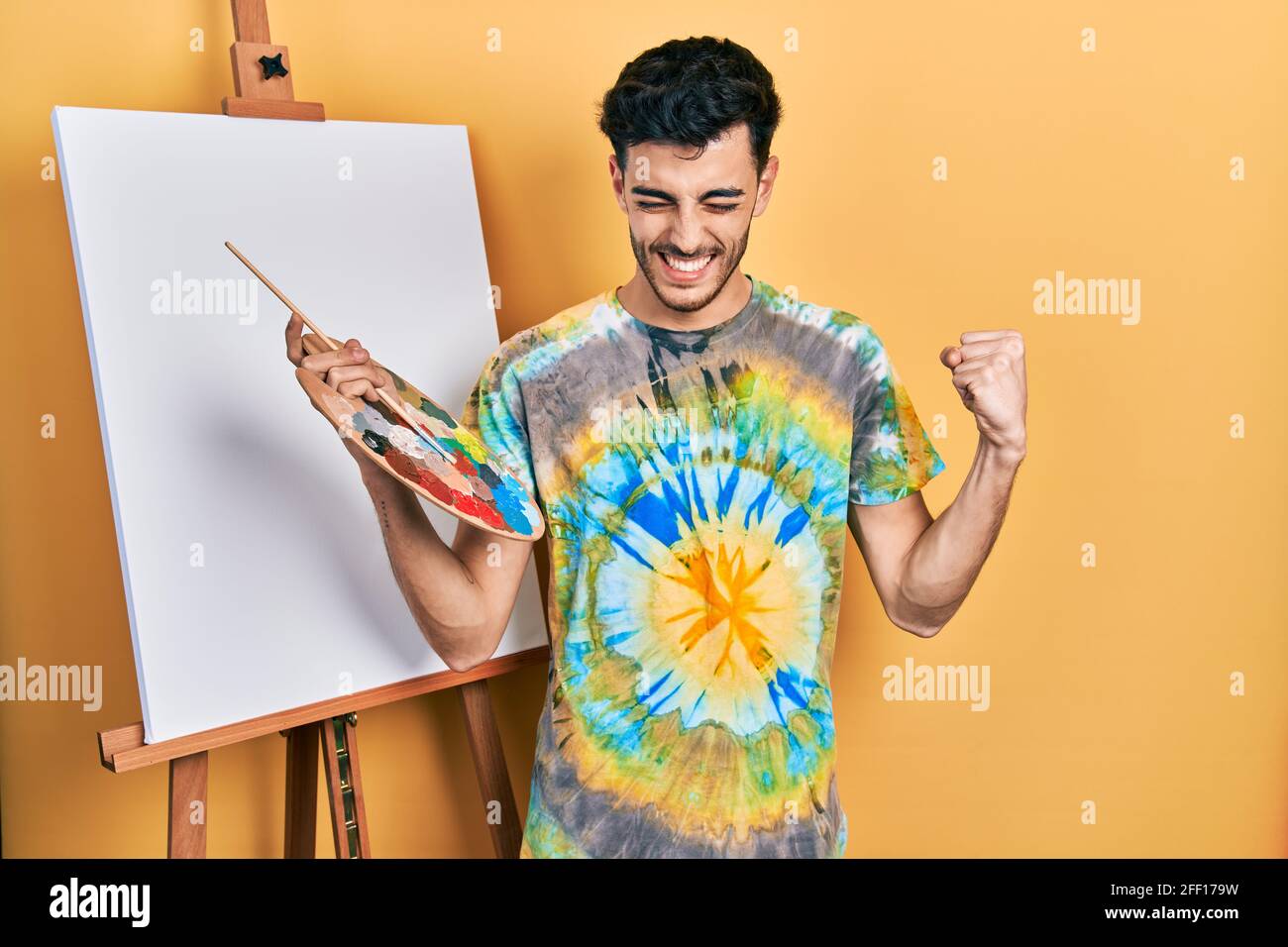 Young hispanic man standing drawing with palette by painter easel stand ...