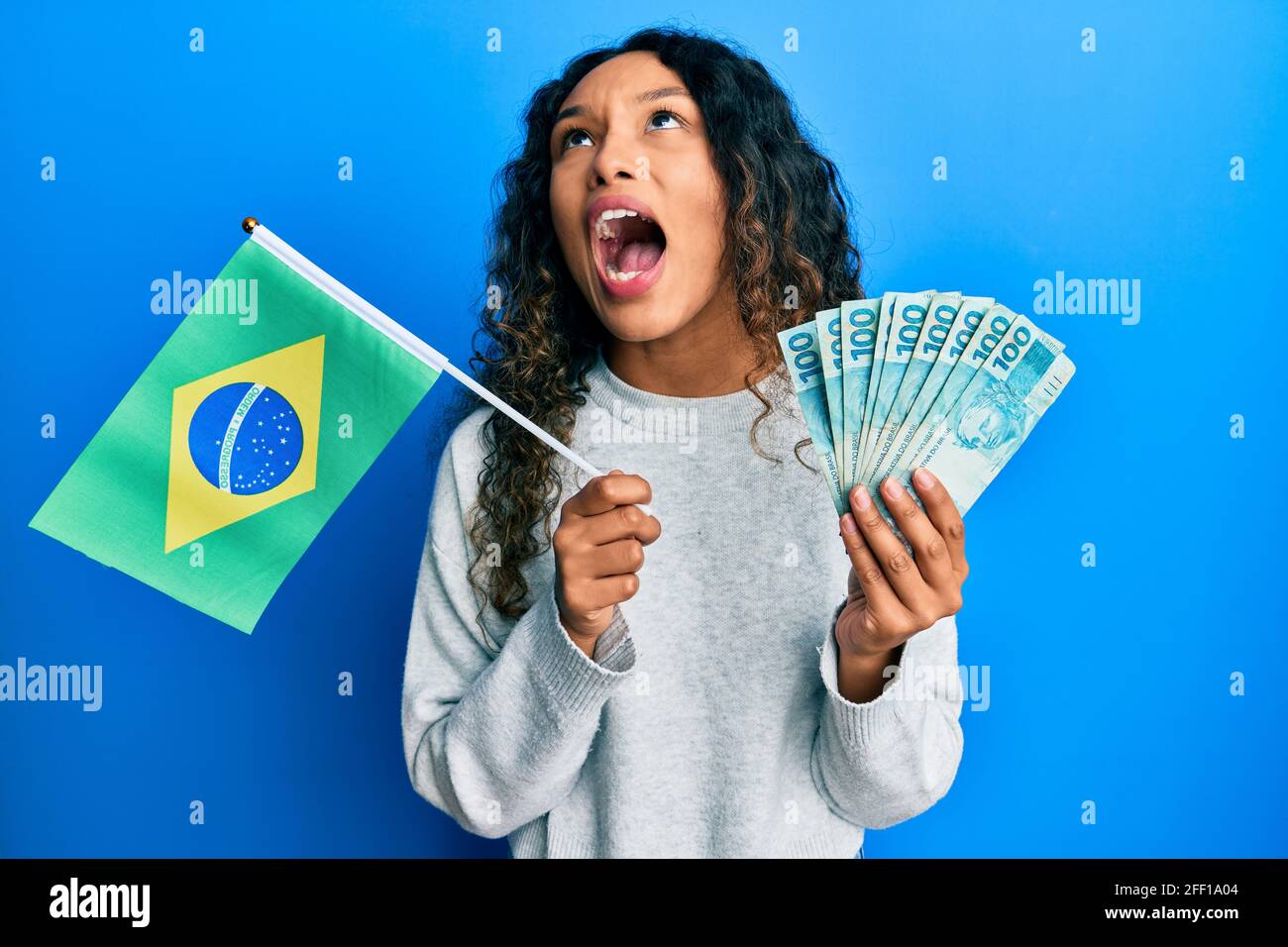 Young latin woman holding brazil flag and real banknotes angry and mad ...