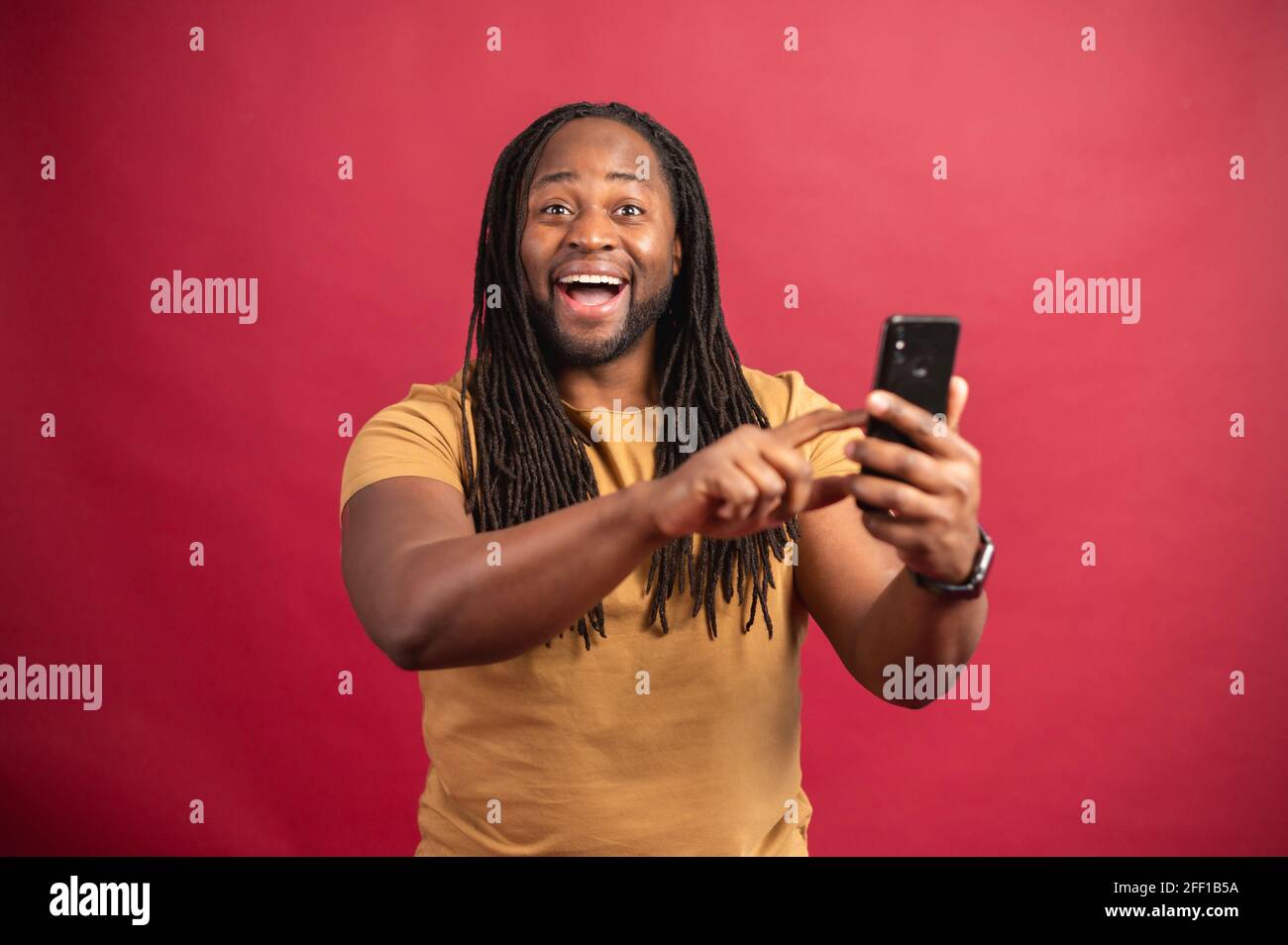 Surprised smiling dark skinned man with long dreadlocks holding phone, amazed African guy ...