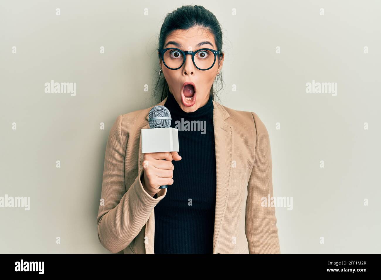 Young hispanic woman holding reporter microphone scared and amazed with ...