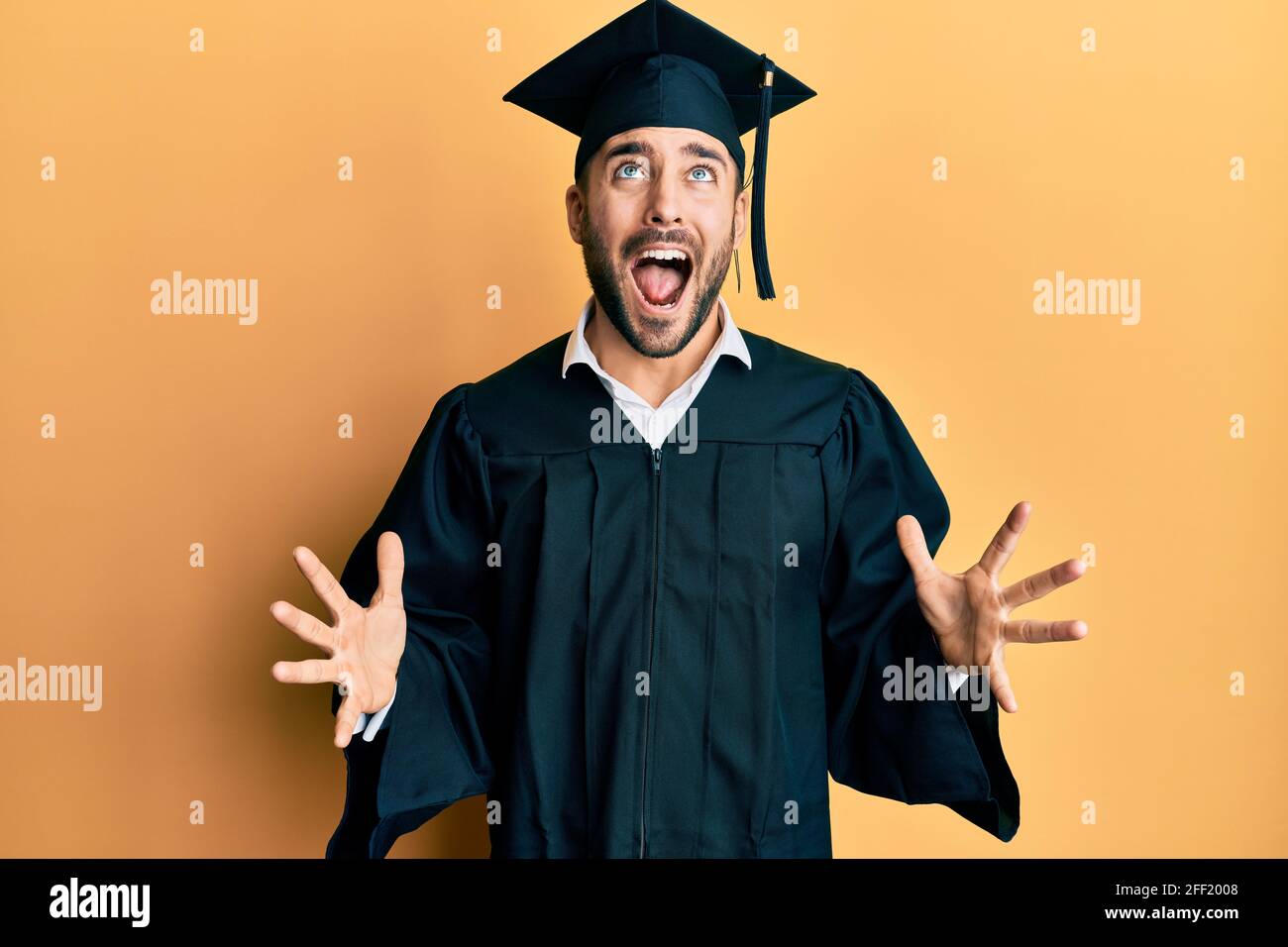Young hispanic man wearing graduation cap and ceremony robe crazy and ...