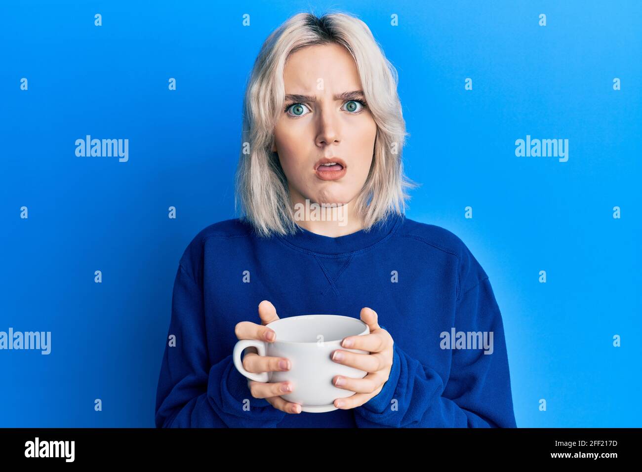 Young blonde girl drinking a cup of coffee clueless and confused ...