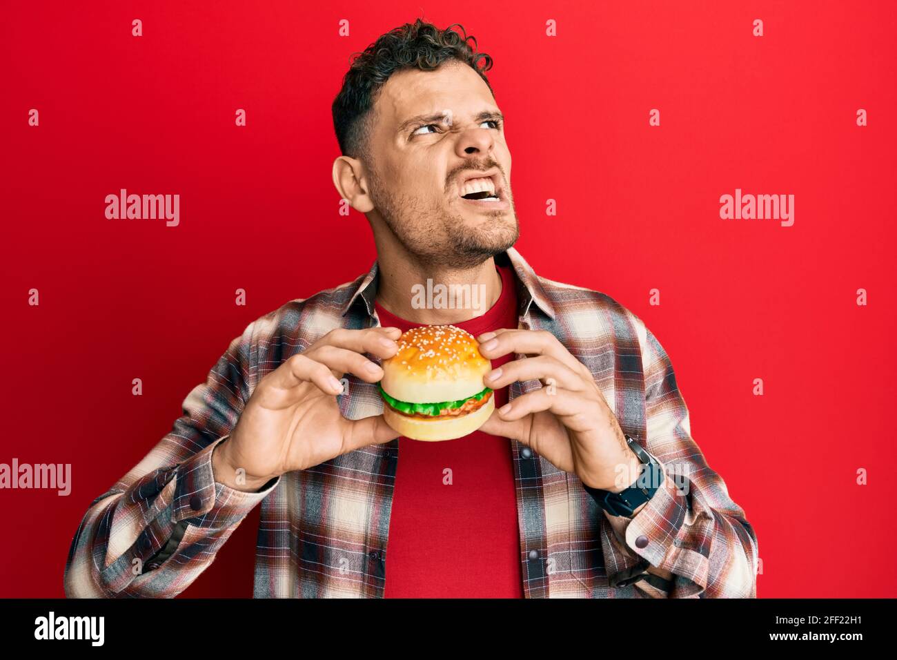 Young hispanic man eating a tasty classic burger angry and mad ...