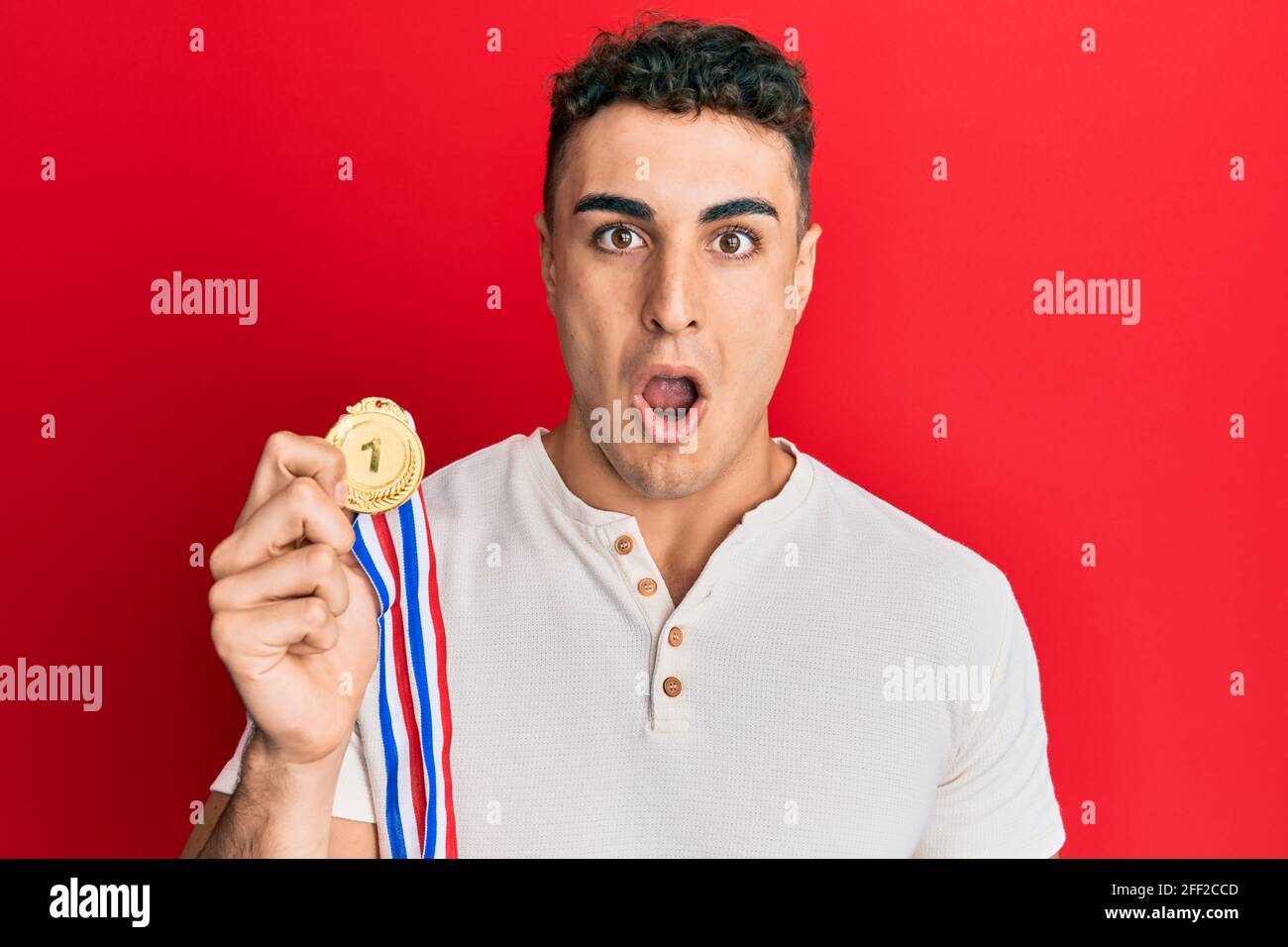 Hispanic young man holding first place medal scared and amazed with ...