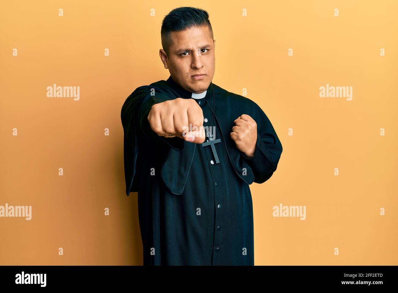 Young latin priest man standing over yellow background punching fist to ...