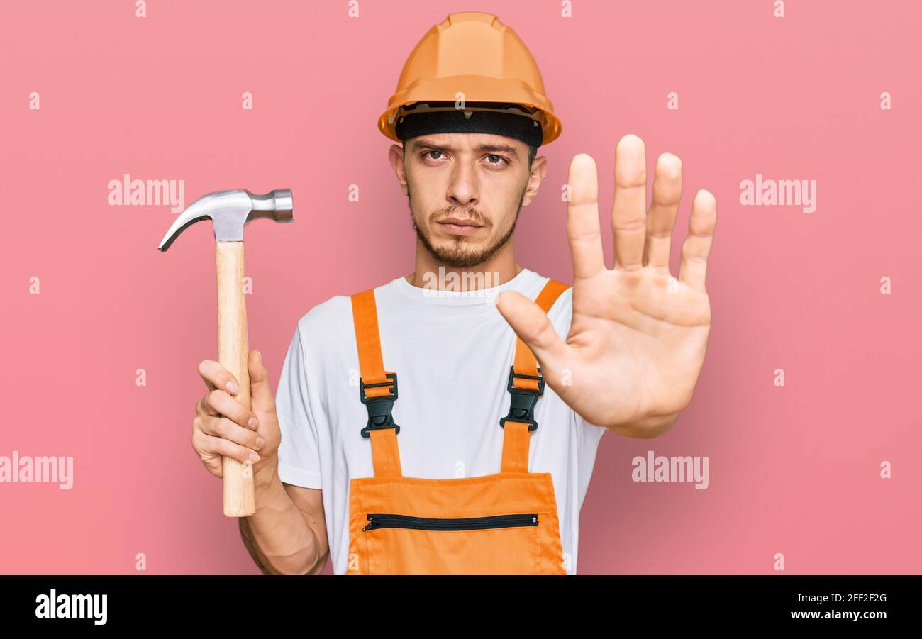 Hispanic young man wearing hardhat holding hammer with open hand doing ...