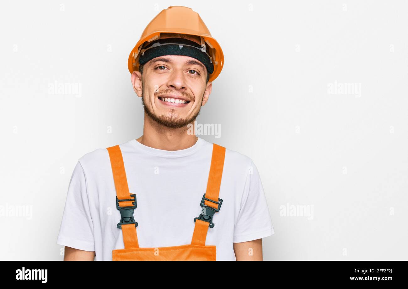Hispanic young man wearing handyman uniform and safety hardhat looking ...