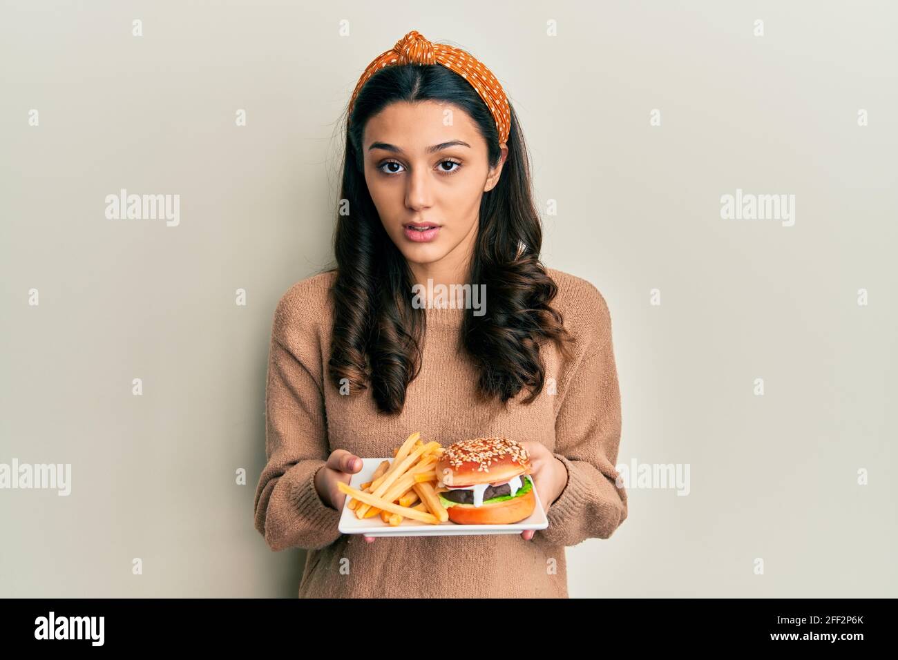 Young hispanic woman eating a tasty classic burger clueless and ...