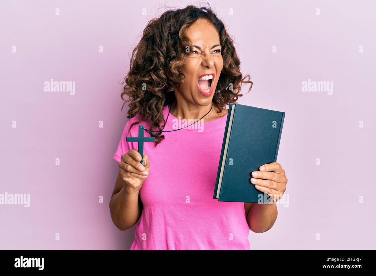 Middle age hispanic woman holding bible and christian cross angry and ...