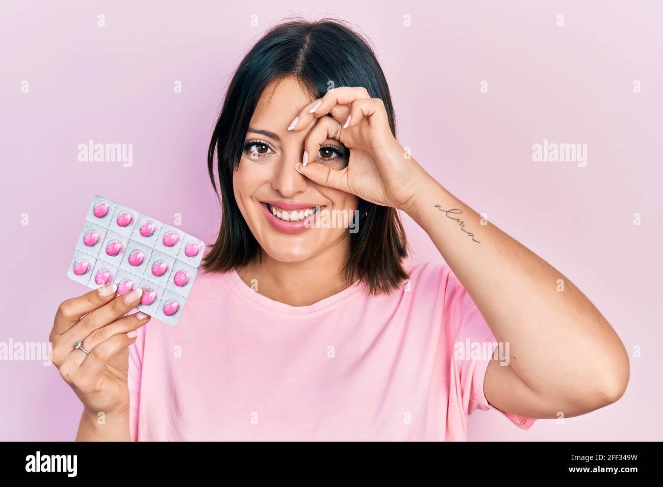Young hispanic woman holding pills smiling happy doing ok sign with ...