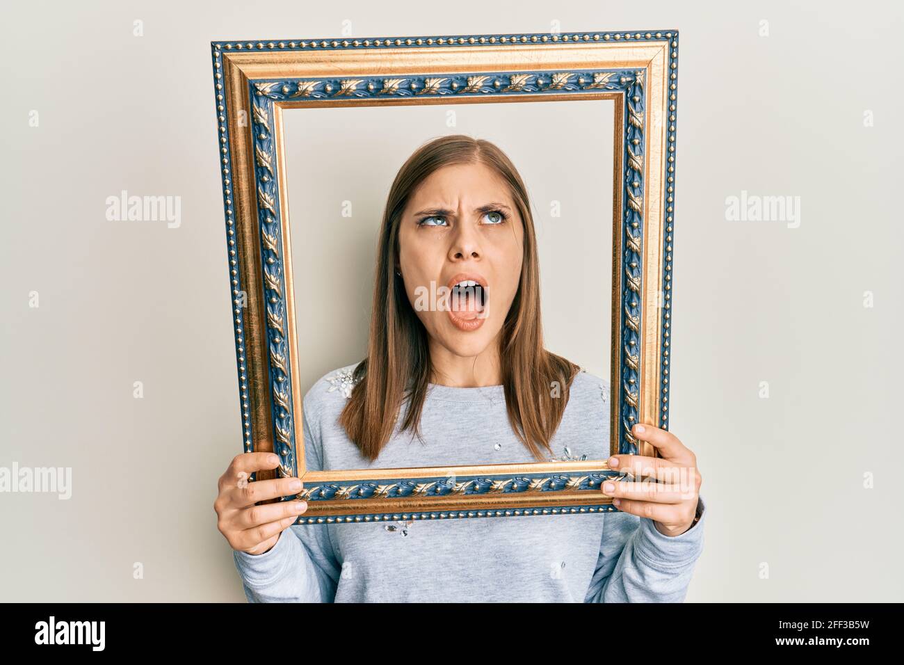 Beautiful caucasian woman holding empty frame angry and mad screaming ...