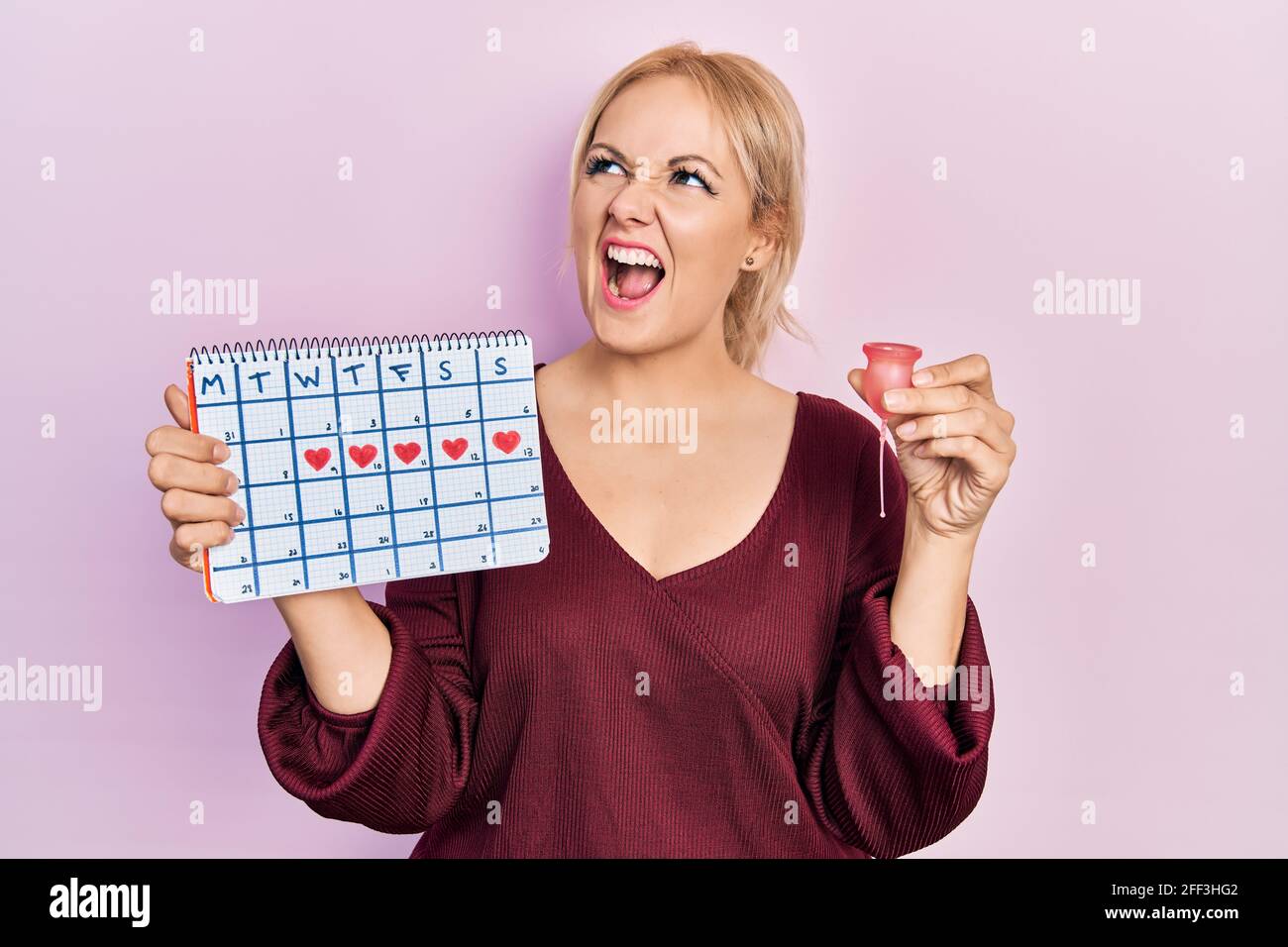 Young blonde woman holding heart calendar and menstrual cup angry and ...
