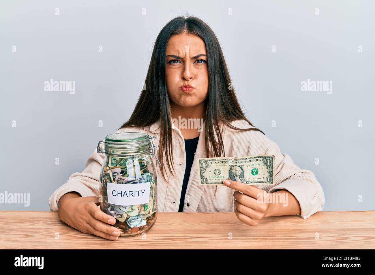 Beautiful hispanic woman holding charity jar with one dollar banknote ...