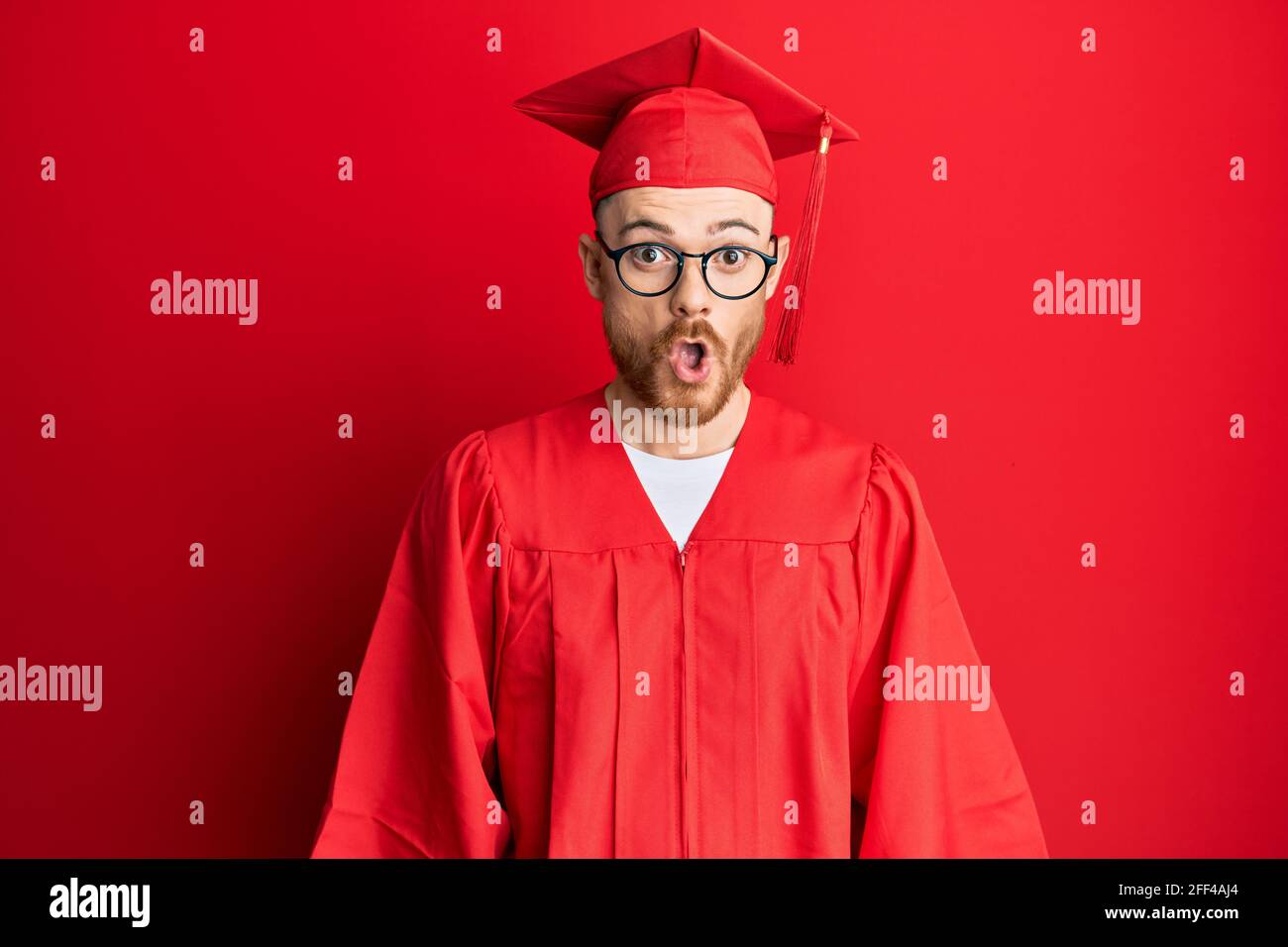 Young redhead man wearing red graduation cap and ceremony robe afraid ...