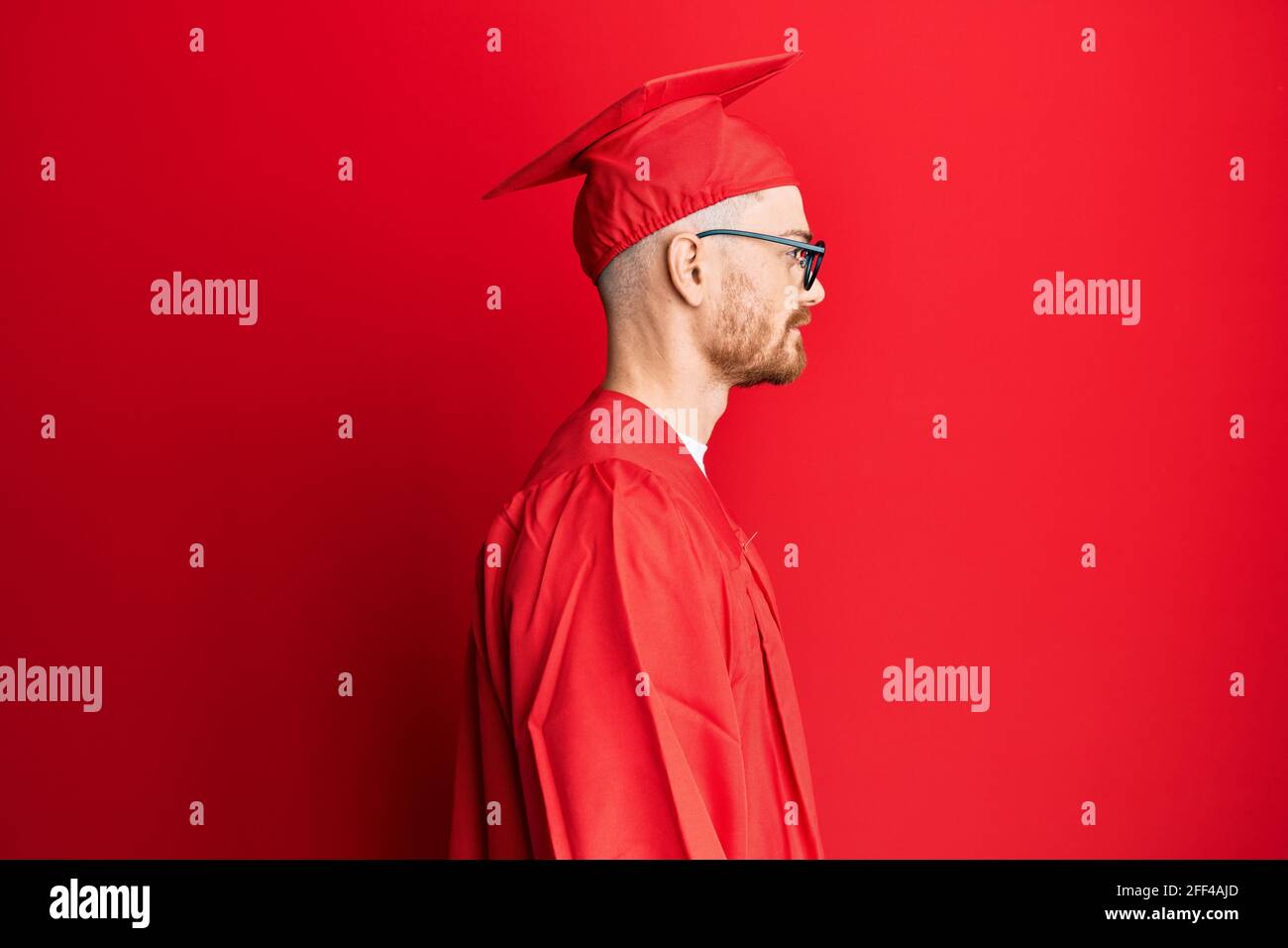 Young redhead man wearing red graduation cap and ceremony robe looking ...