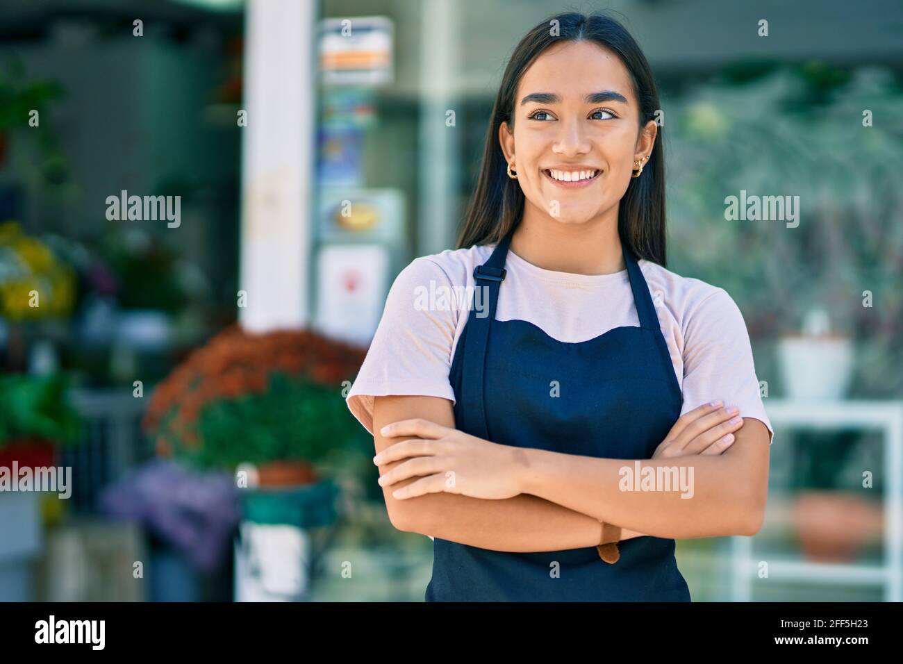 Young latin shopkeeper girl with arms crossed smiling happy standing at ...