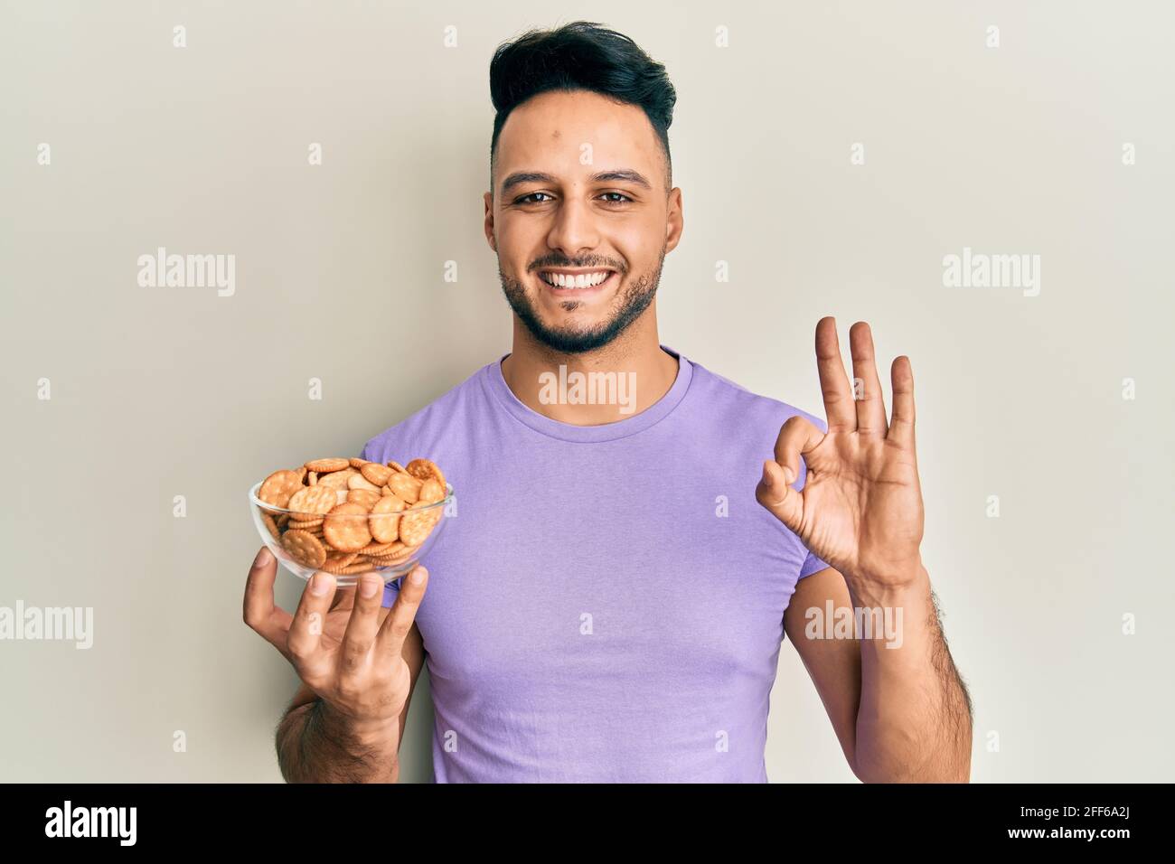 Young arab man holding bowl of salty crackers biscuits doing ok sign ...