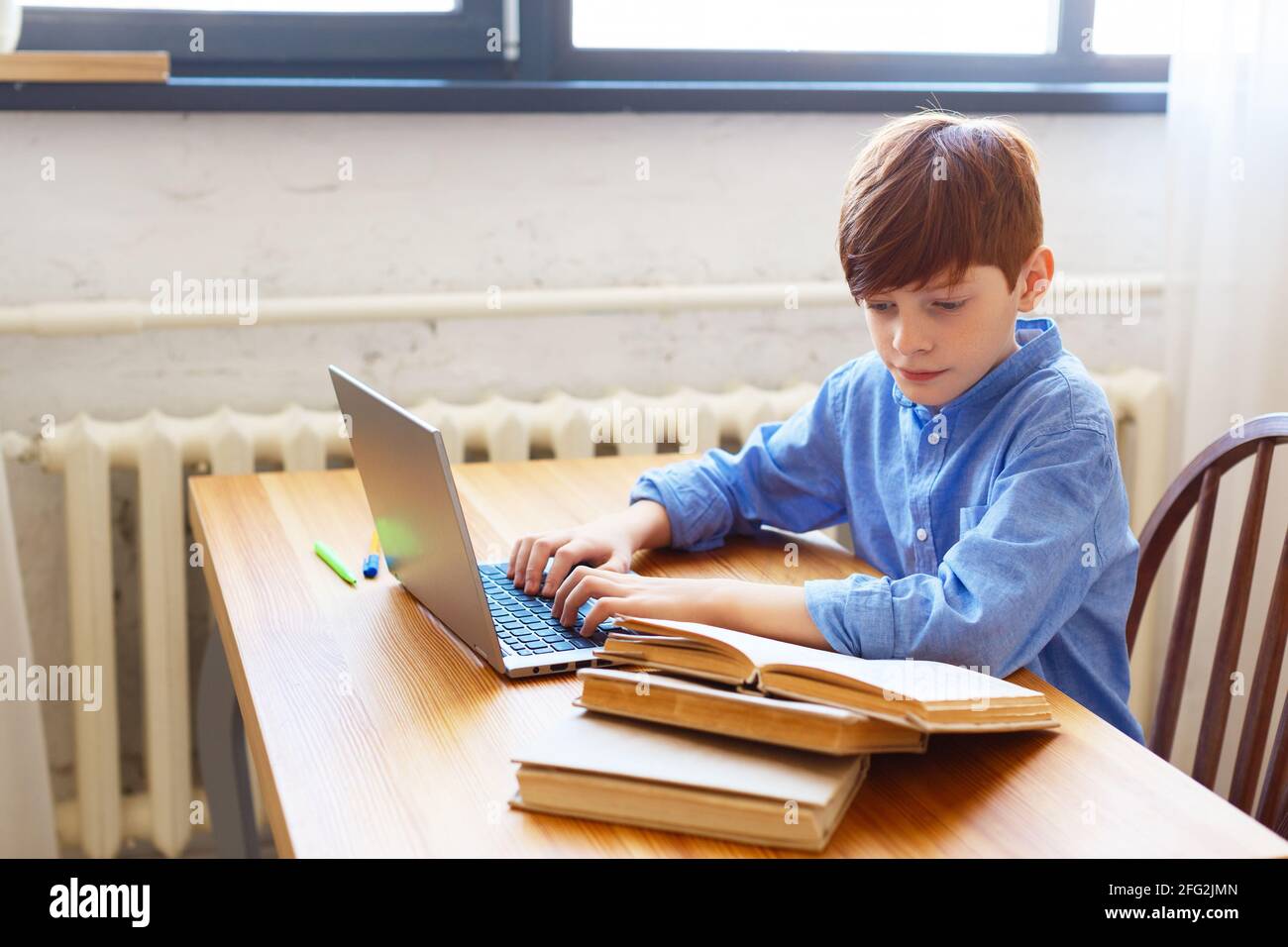 Teen boy doing homework online at home. He looks at the textbook and ...