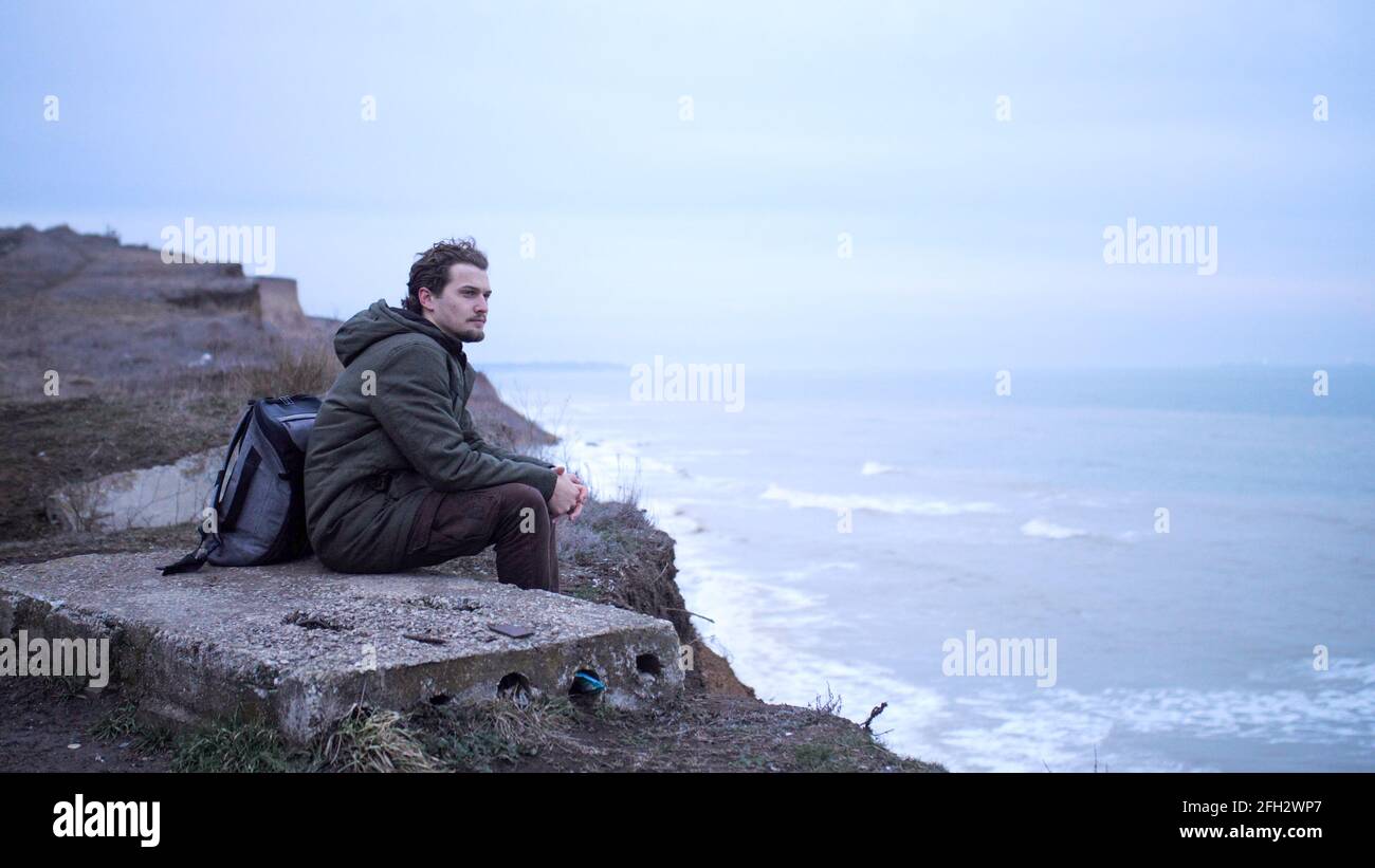 Sad lonely young man sits on a cement block at the edge of cliff near ...