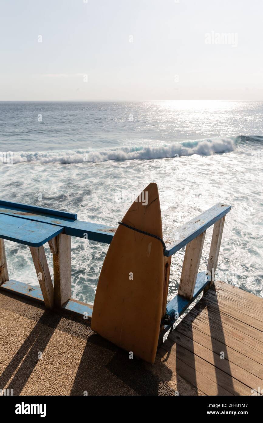 Surfboard placed on wooden quay near wavy foamy sea under clear blue ...