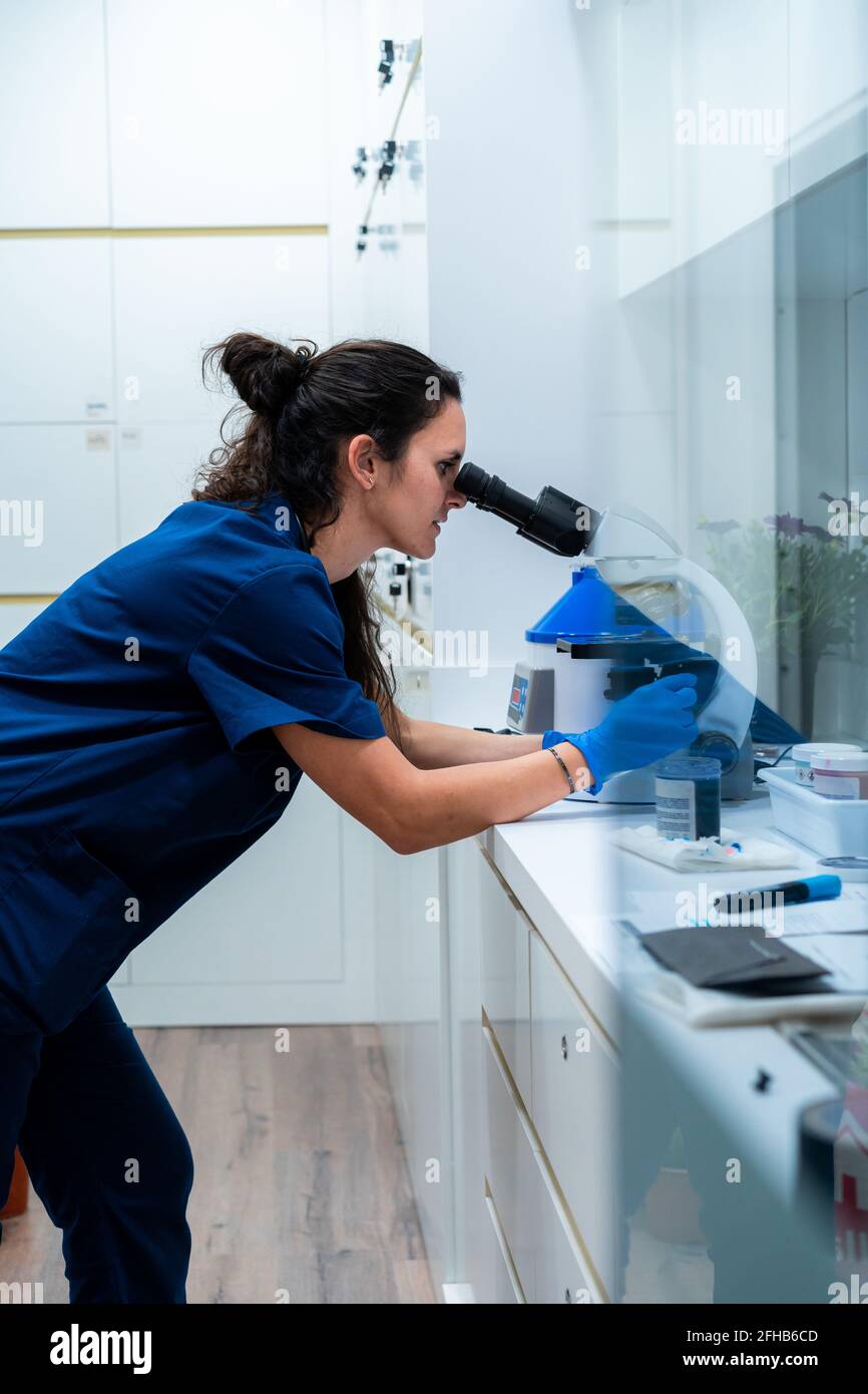 Side view of female vet in blue uniform looking through microscope ...