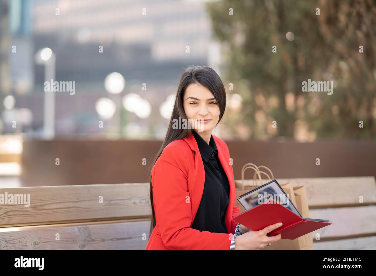 Smiling female customer sitting on bench with paper bags and reading ...