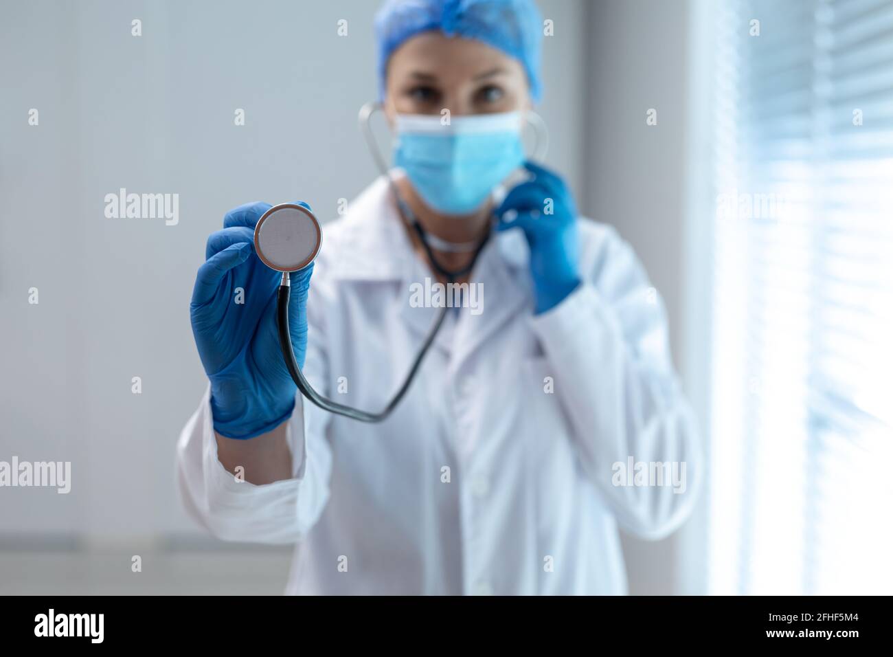 Caucasian female doctor wearing face mask holding stethoscope to camera ...