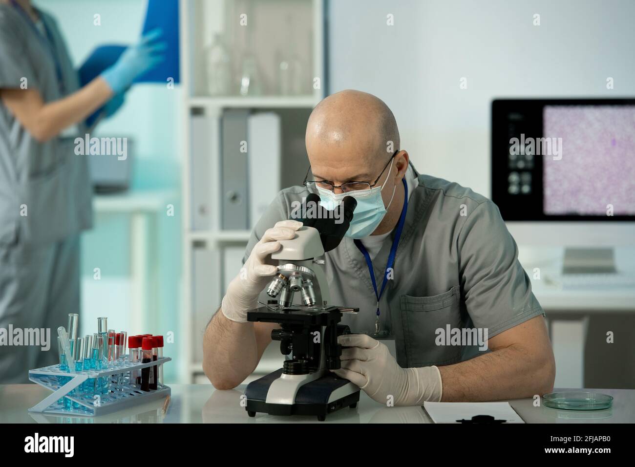 Concentrated bald lab worker scrubs and gloves sitting at table and ...