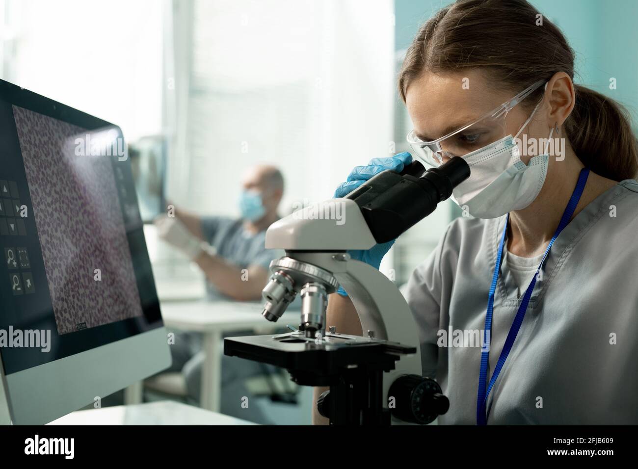 Concentrated young lab researcher in goggles and mask sitting in front ...