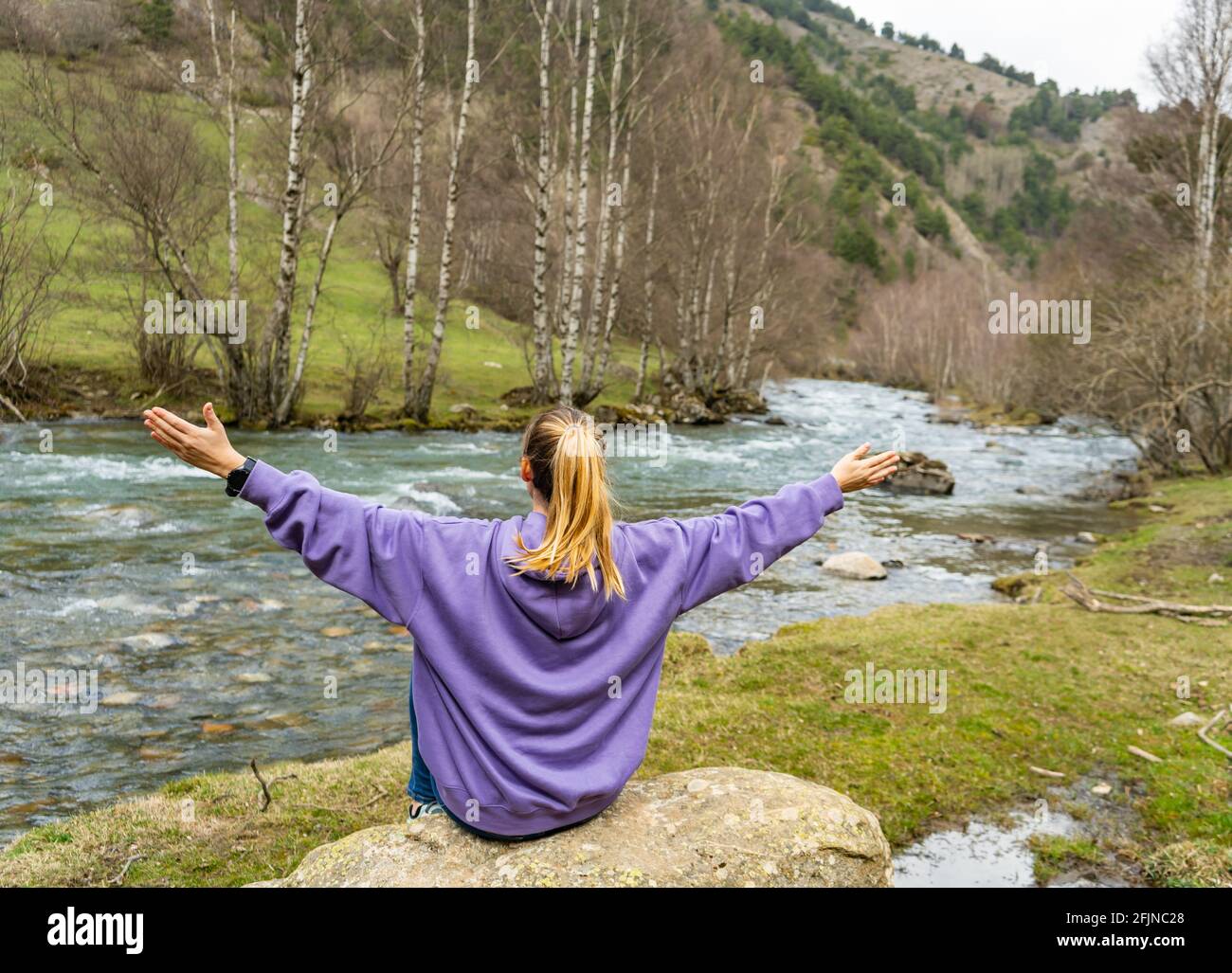 Happy woman with raised up arms sitting on a rock near to the river ...