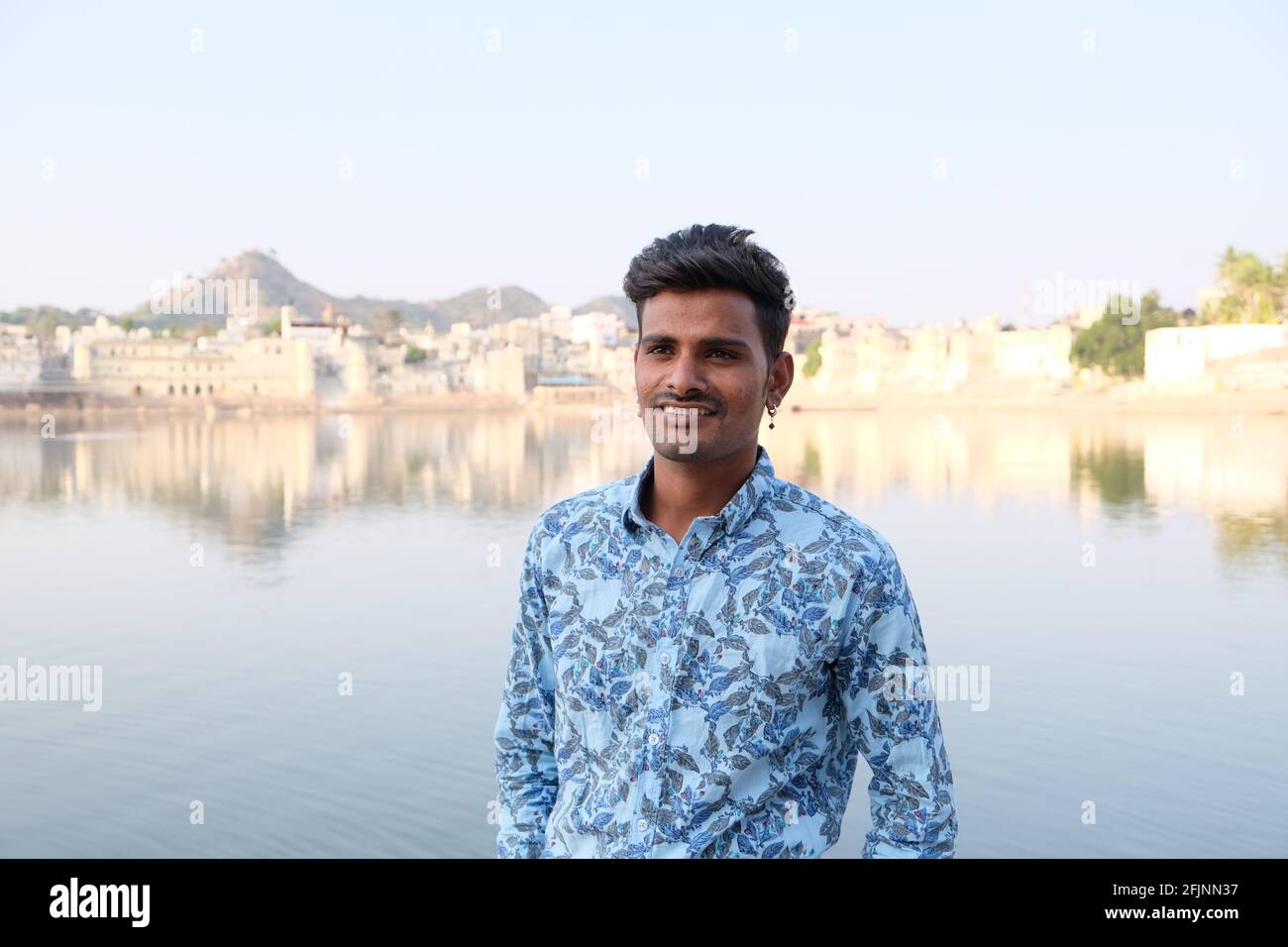 A portrait of a young smiling Indian male posing outdoors with a lake ...