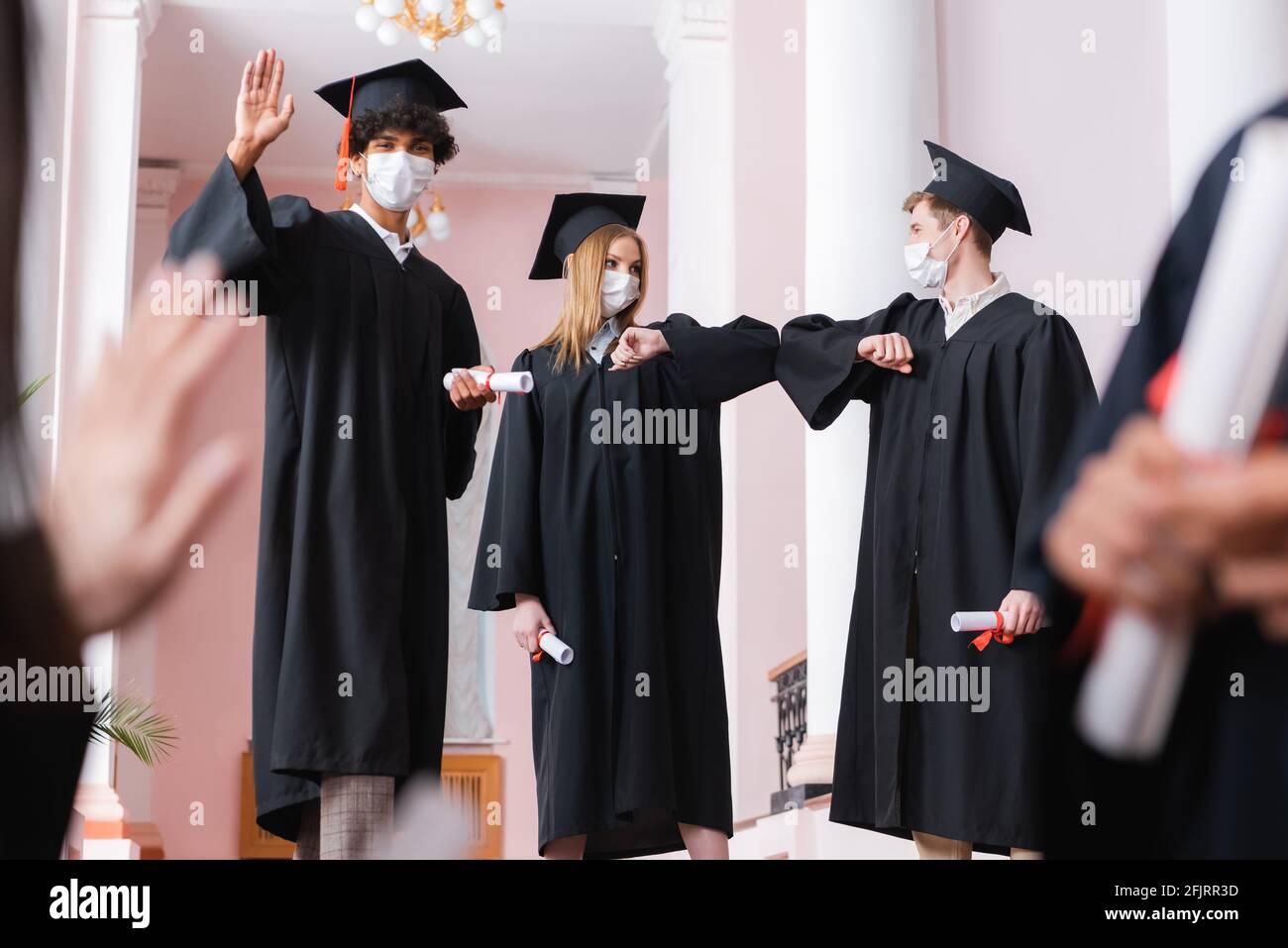 African american graduate in medical mask waving hand near friends ...