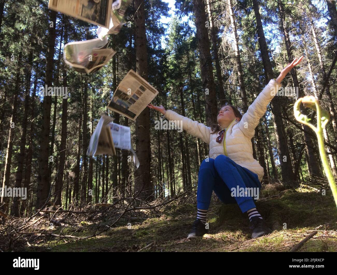 A girl sitting in a forest throwing a newspaper into the air with her ...