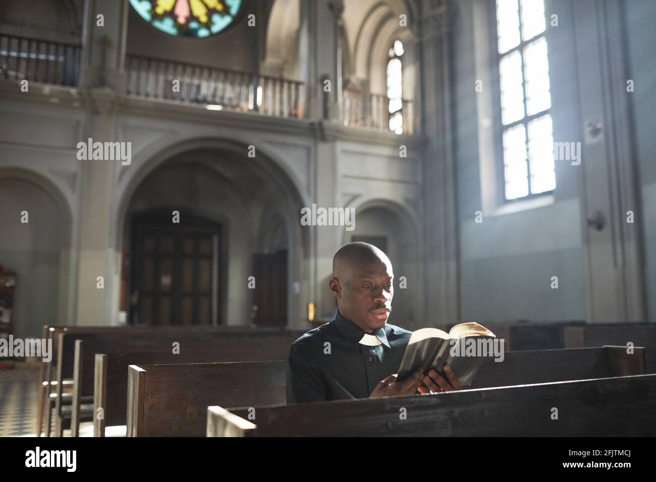 African priest in collar sitting on the bench in church and reading the ...