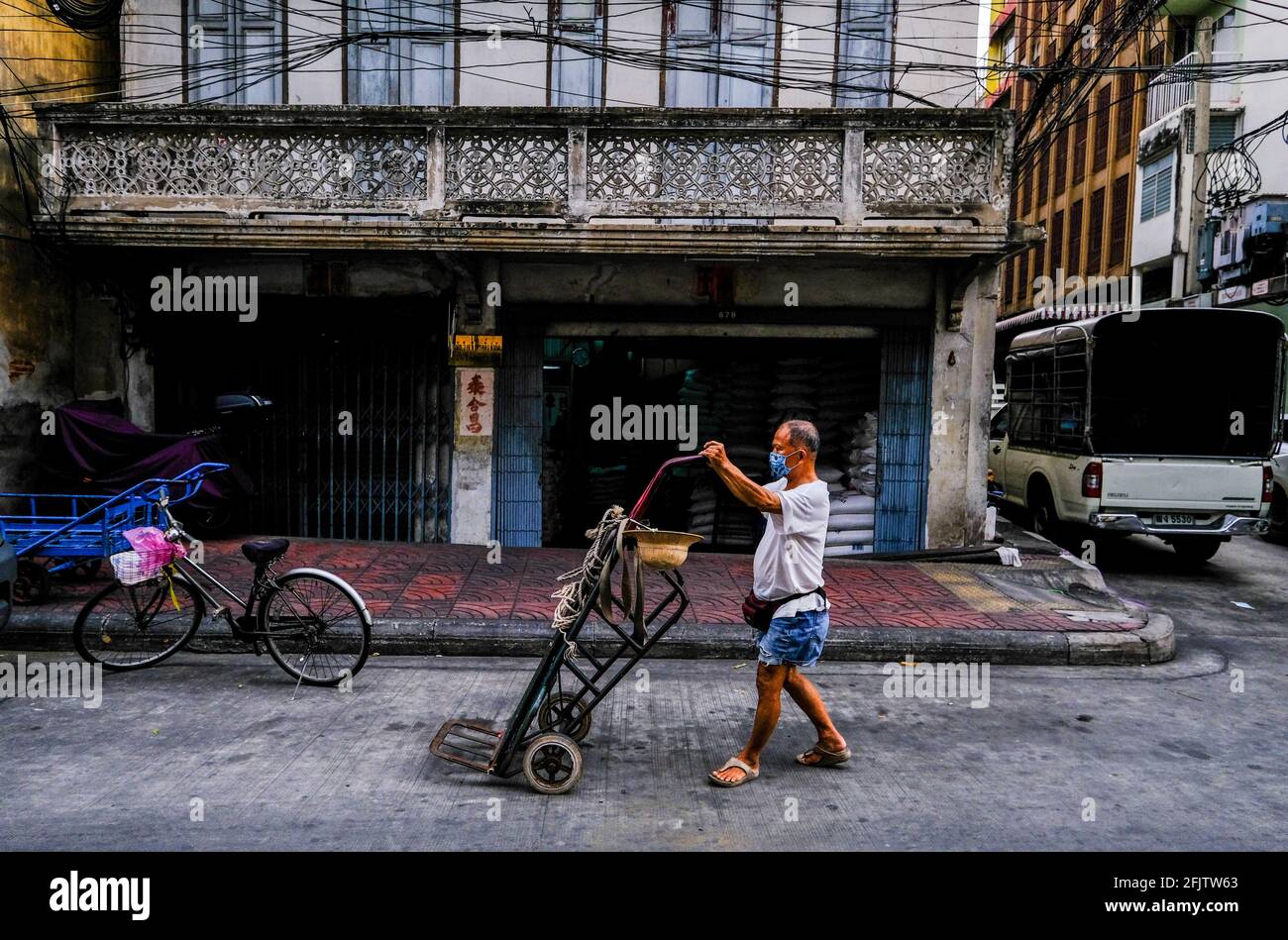 An elderly Thai male porter pushes an empty trolley along a street in ...
