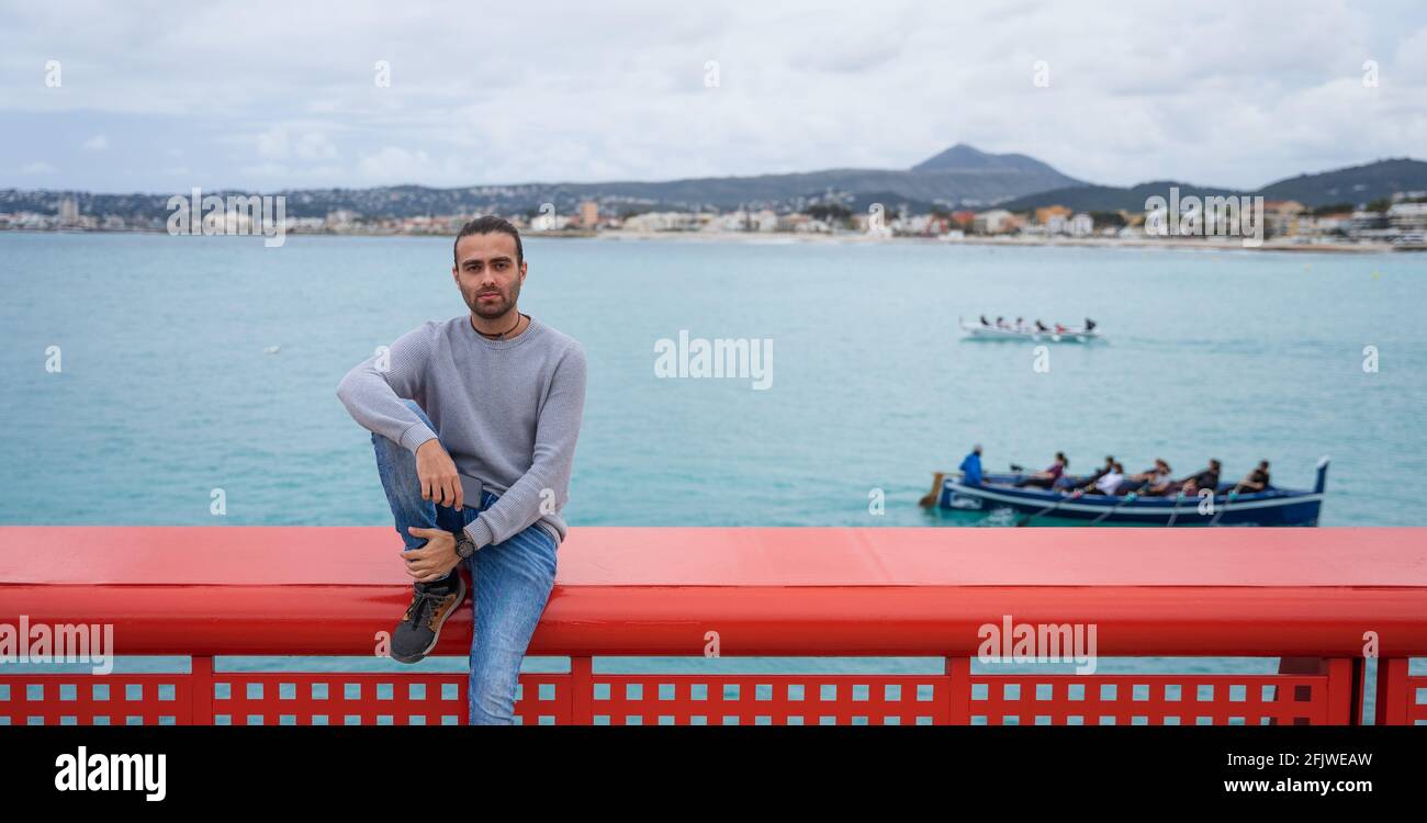 Man sitting on a red railing. He is on a promenade, on a cloudy day. He ...