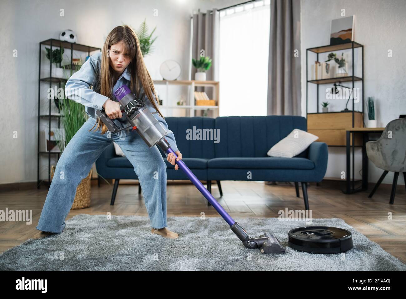 Playful young woman in denim clothes cleaning her modern apartment with ...