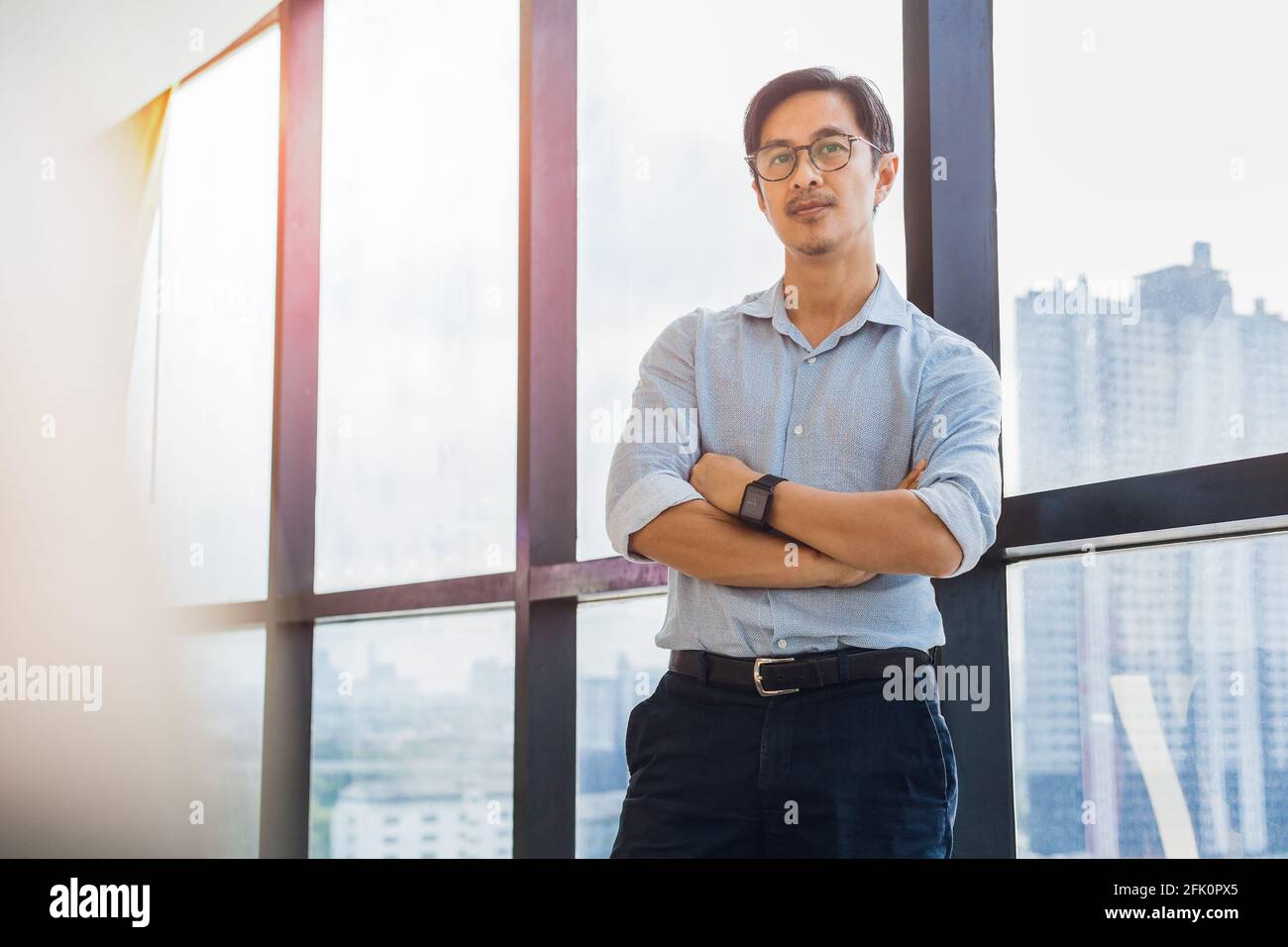 Portrait of business man standing next to office window with arms ...
