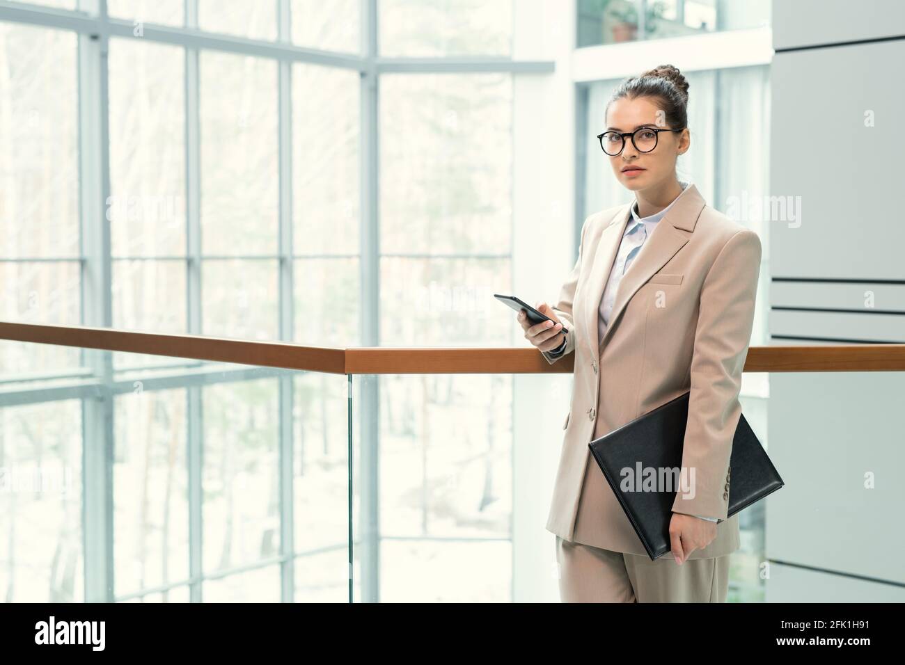 Portrait of serious young female manager in beige costume standing with ...