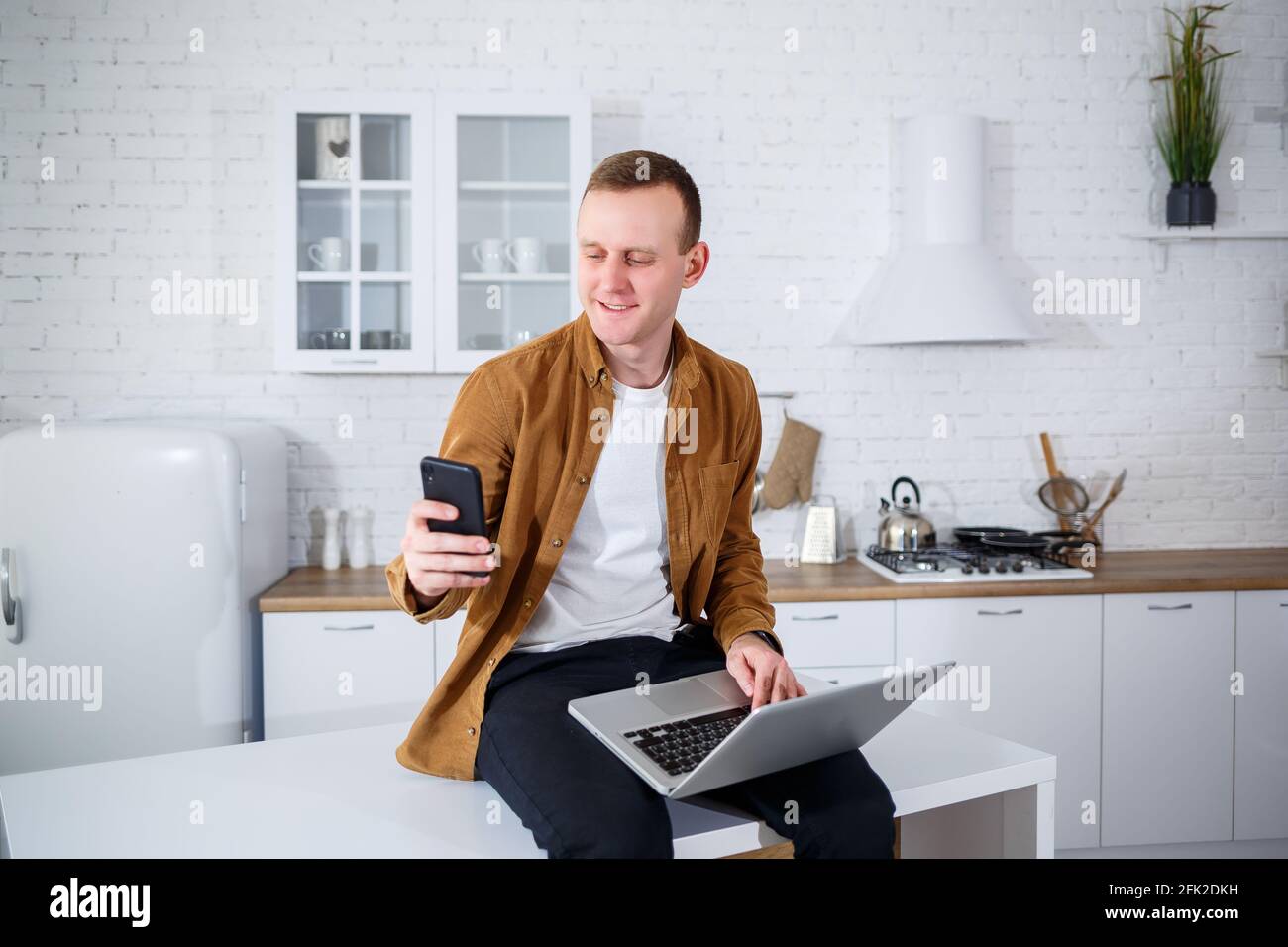 An attractive young man in casual clothes sitting in the kitchen using ...