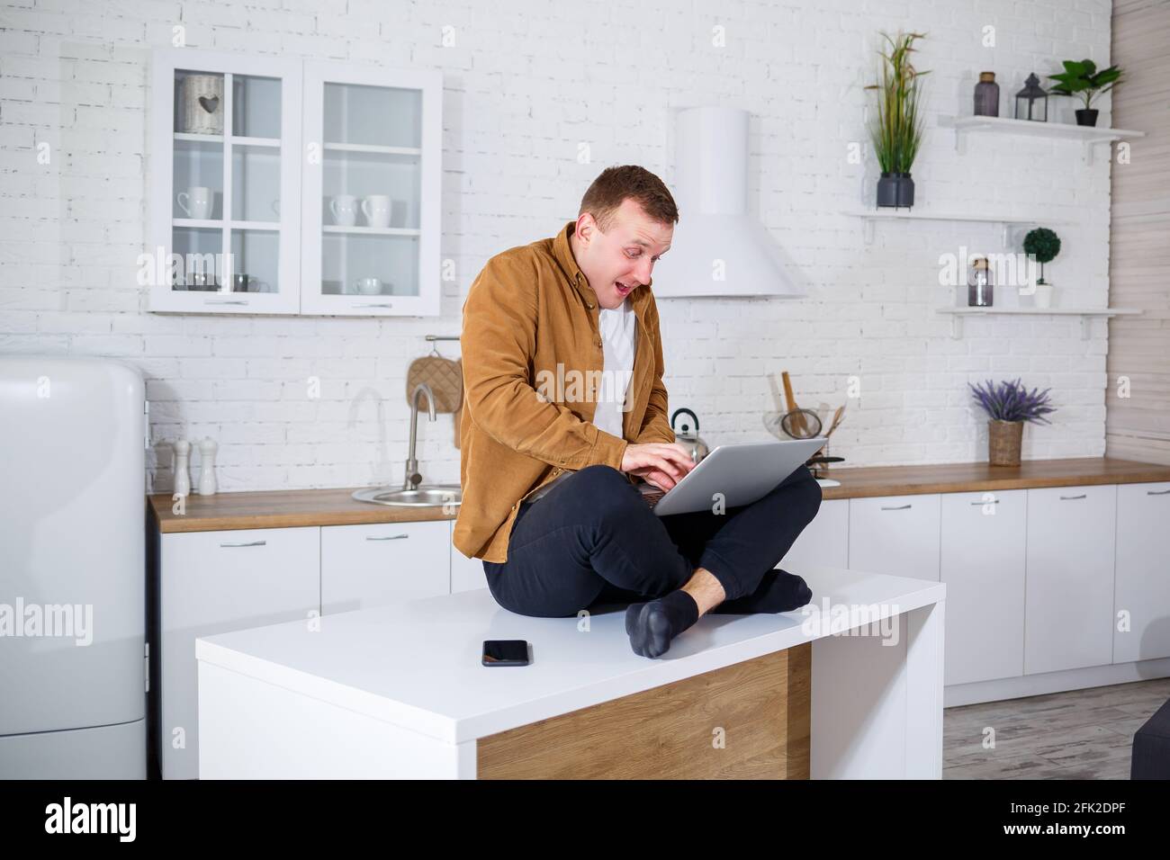 An attractive young man in casual clothes sitting in the kitchen using ...