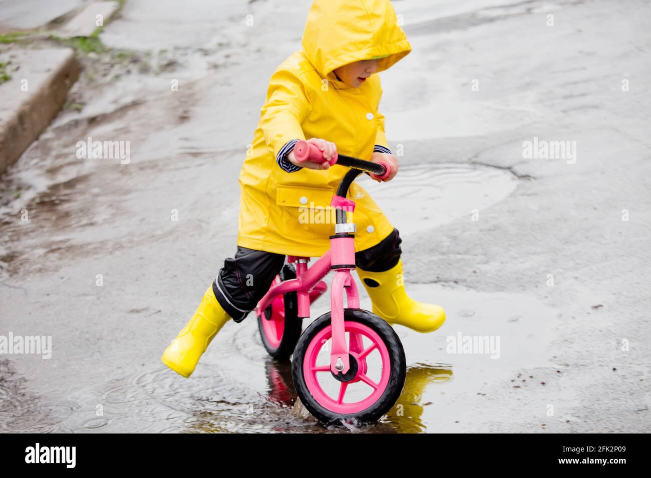 little girl in yellow raincoat rides on bike ride through puddles in ...