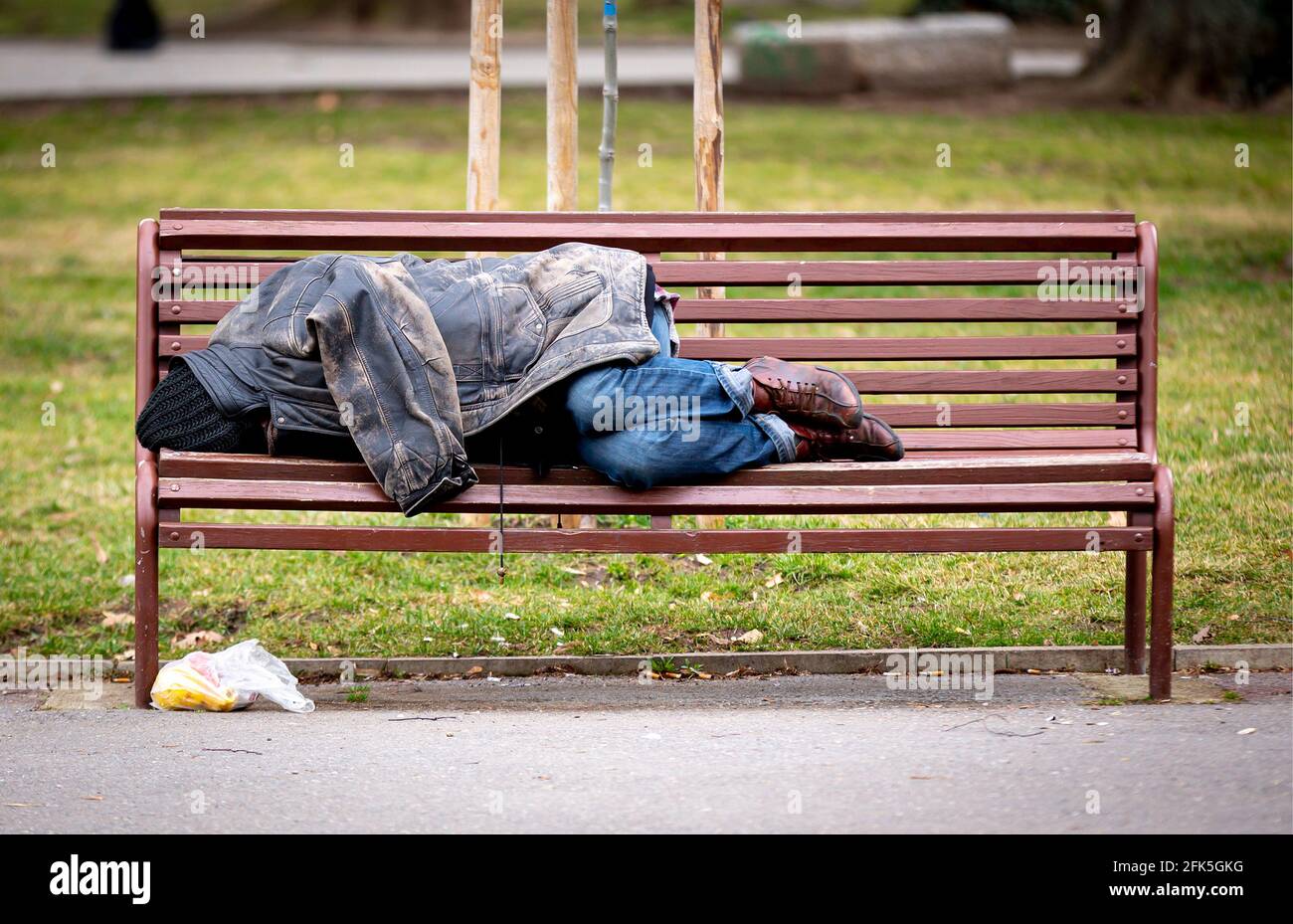 Homeless man sleeps on a bench covered with his jacket in a public park ...