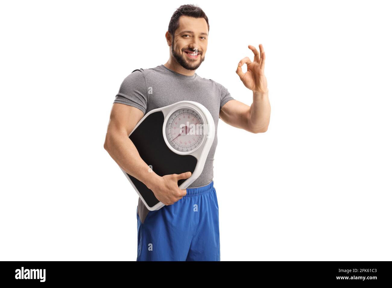 Smiling young man holding a weight scale and gesturing ok sign isolated ...