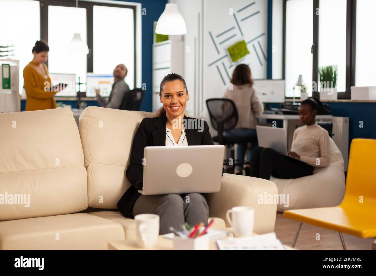 Hispanic business woman smiling at camera sitting on couch typing on ...