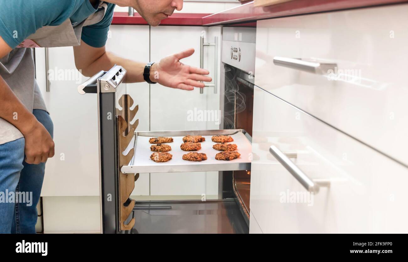 A young man taking homemade cookies out of the oven and appreciating ...
