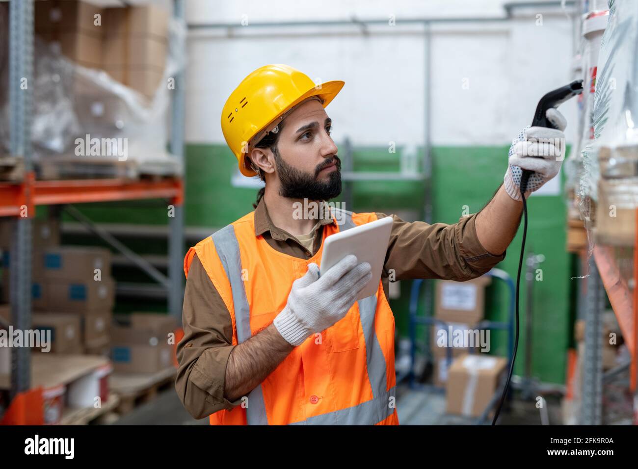 Serious young bearded man in hardhat and gloves scanning code of item ...