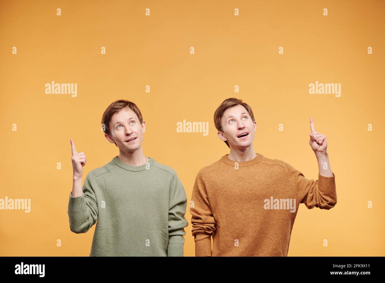 Surprised young twins in colorful sweaters standing against orange ...
