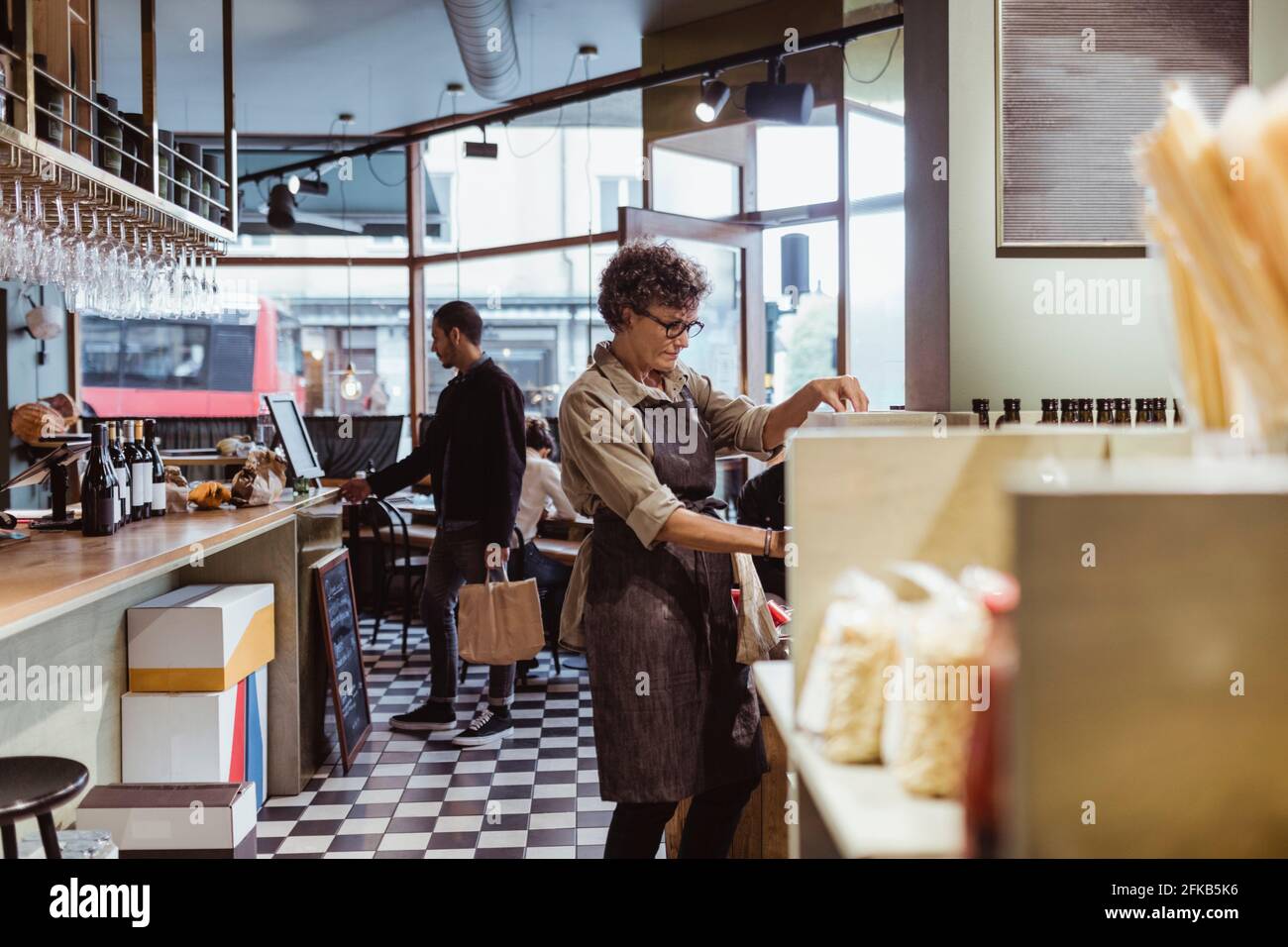 Female store owner working in deli store with customers in background ...