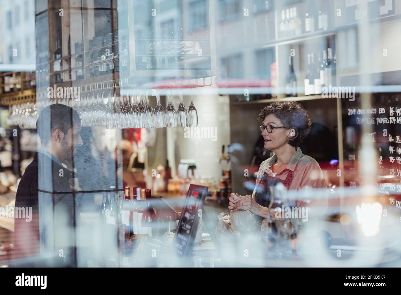 Male customer collecting order from female owner seen through glass in ...